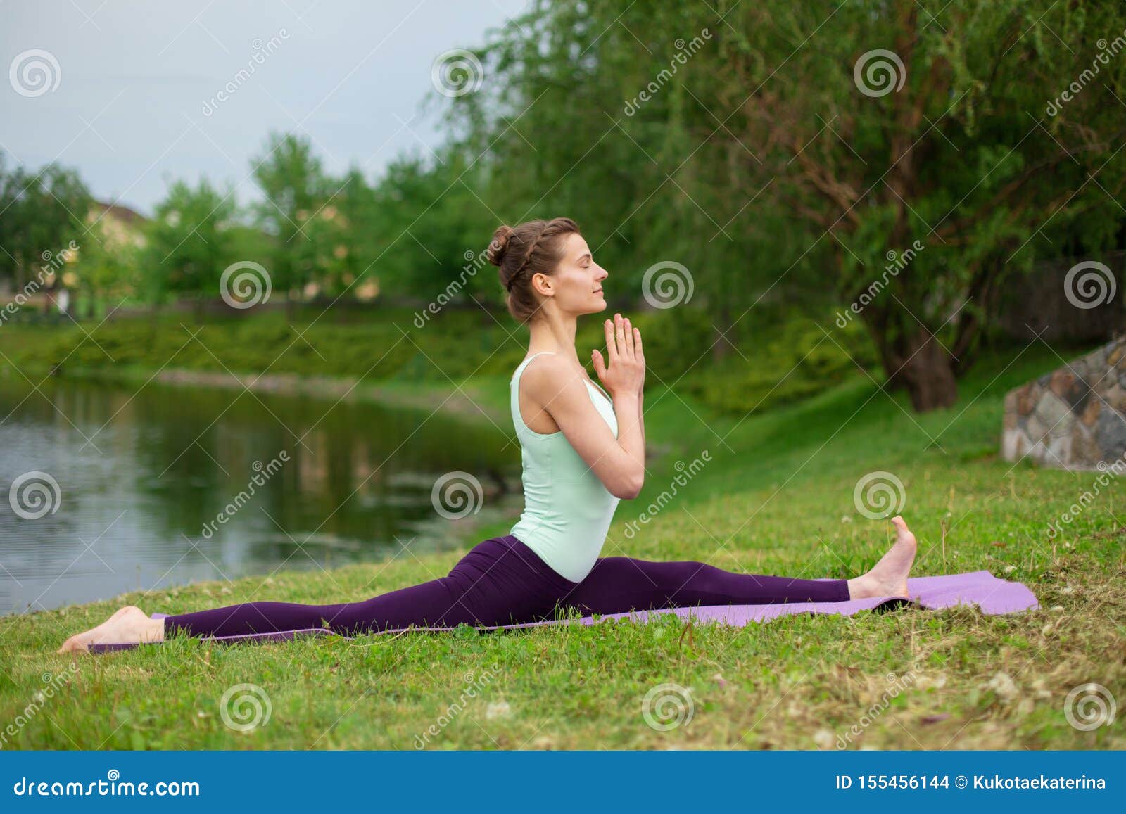 Slim Brunette Girl Doing Yoga In The Summer On A Green Lawn By The Lake