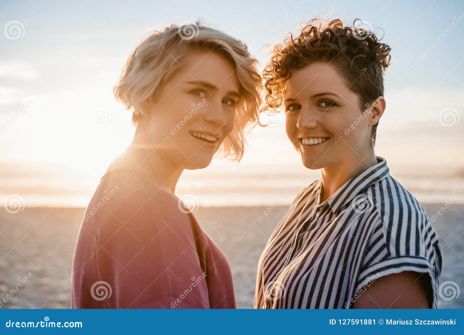 Smiling Lesbian Couple Standing Together On A Beach At Sunset Stock