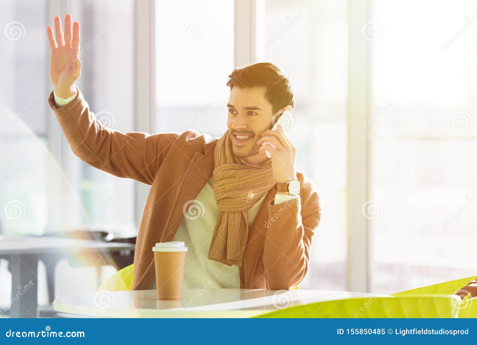 Smiling Man Sitting at Table, Talking on Smartphone and Waving with ...