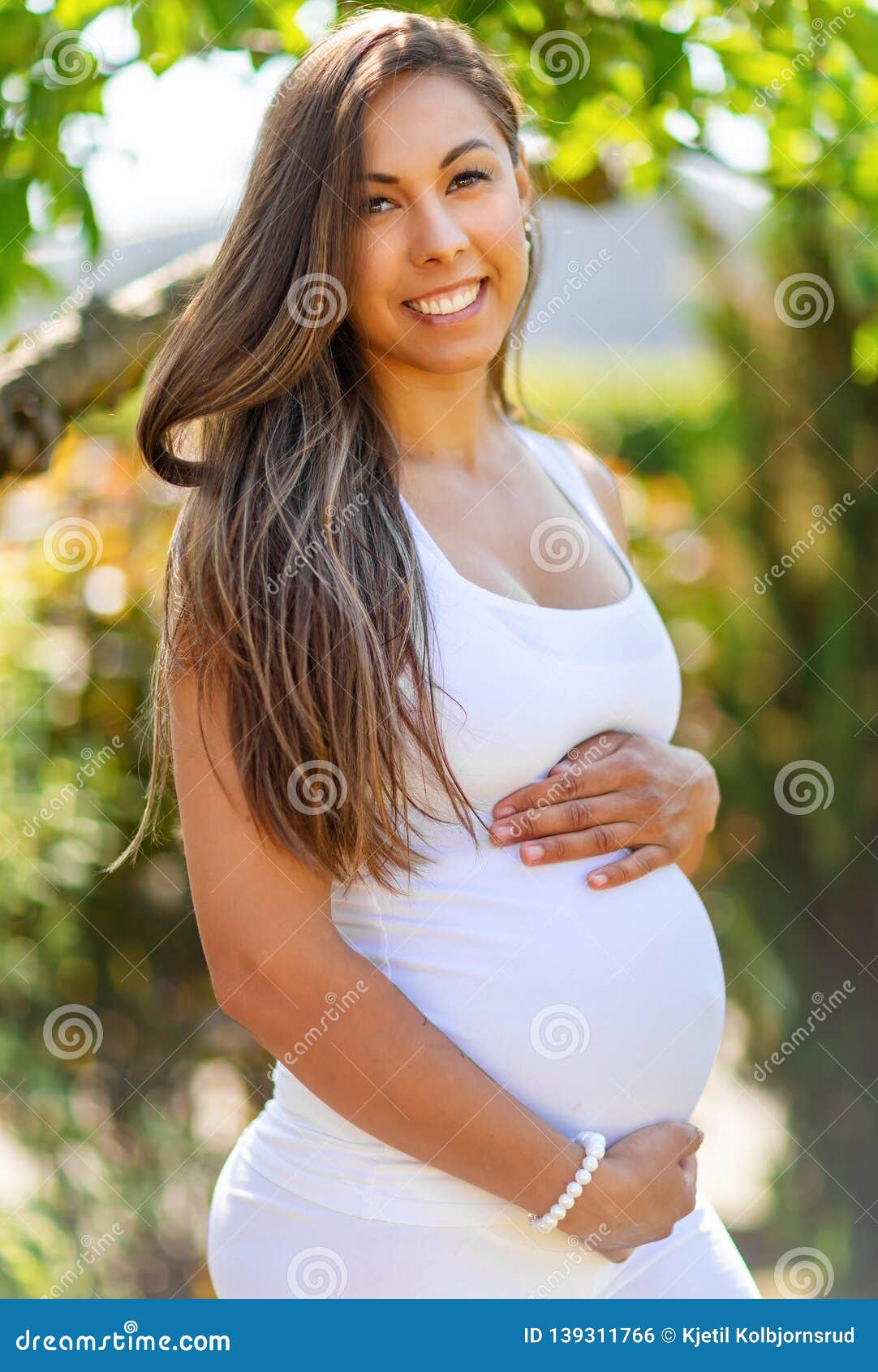 Smiling Pregnant Woman Standing In Garden Holding Hands On Belly Stock