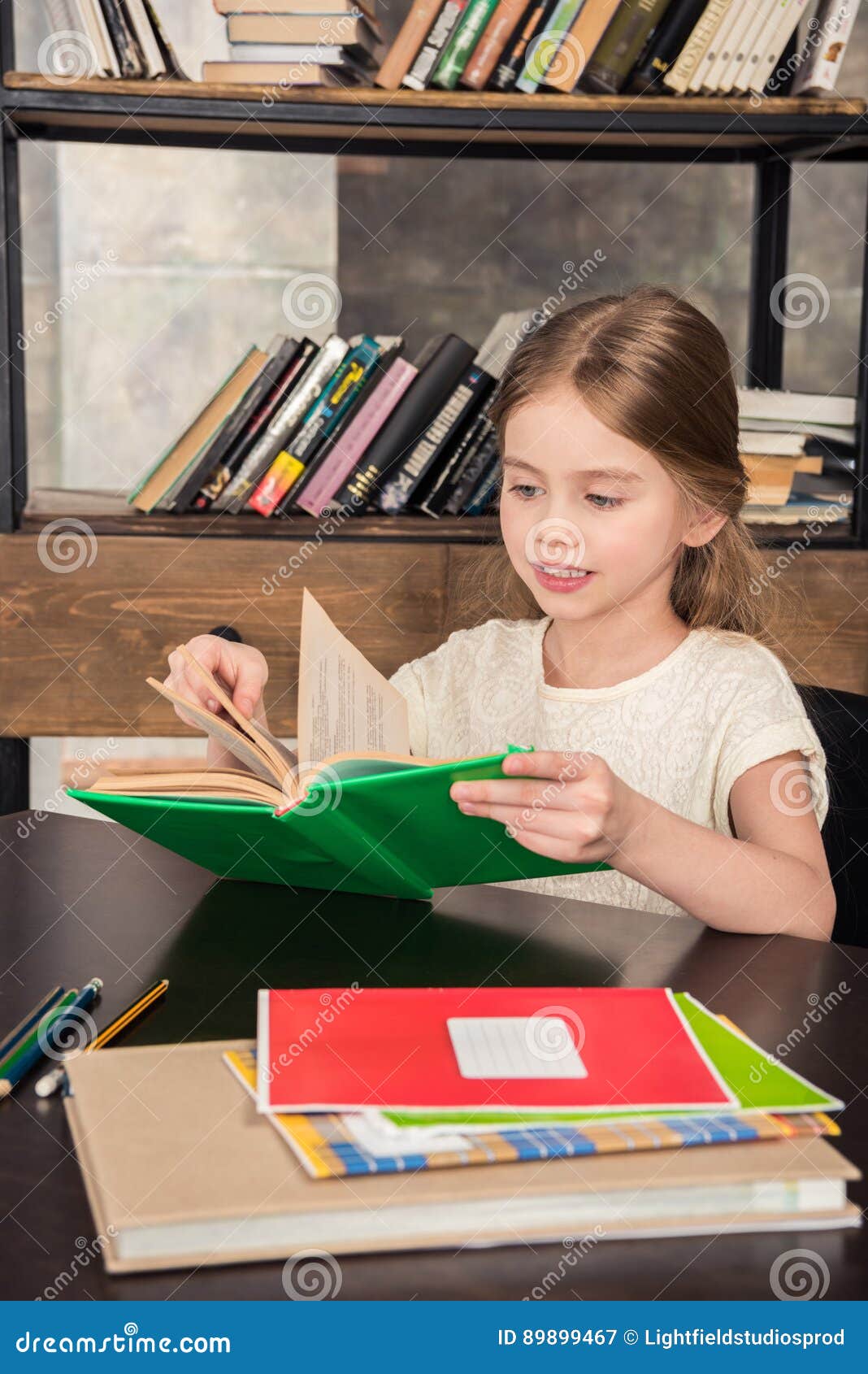 Smiling Schoolgirl Sitting At Table And Reading Book In Library Stock ...