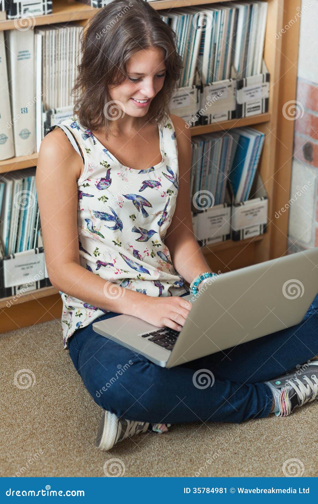 Smiling Student Against Bookshelf Using Laptop on the Library Floor ...