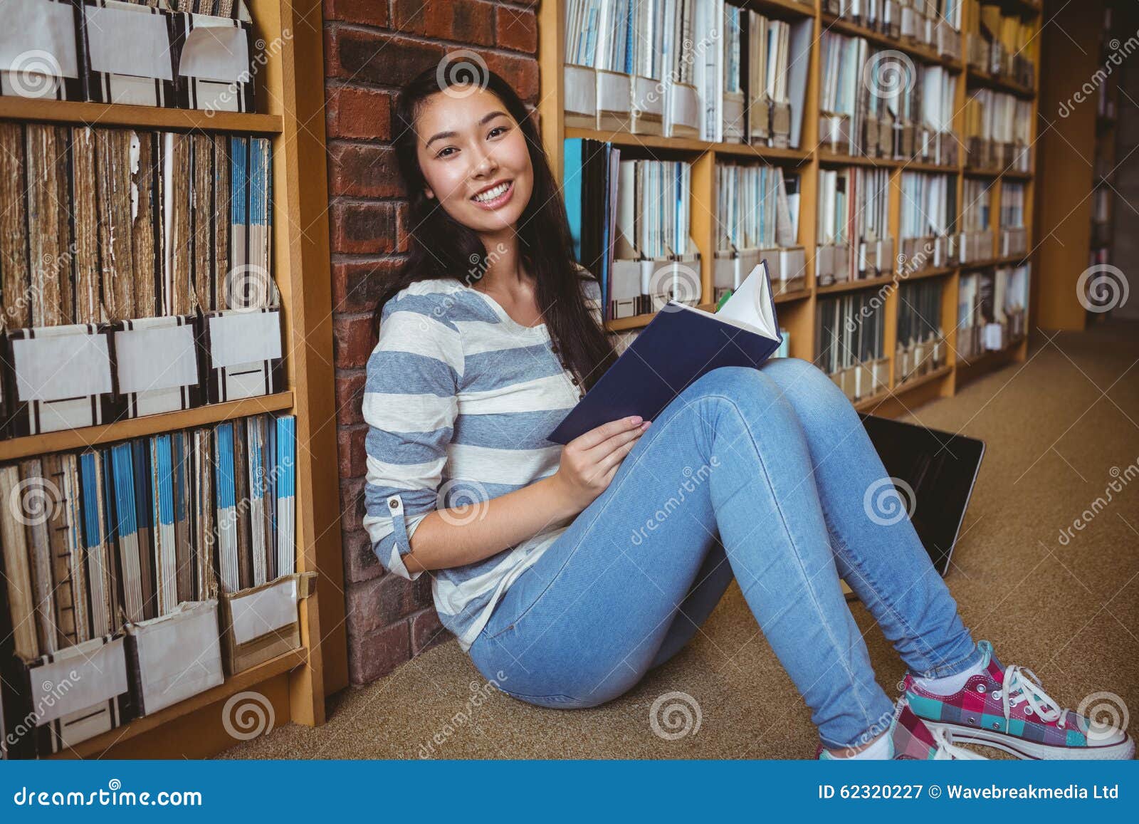Smiling Student Sitting on the Floor Against Wall in Library Reading ...