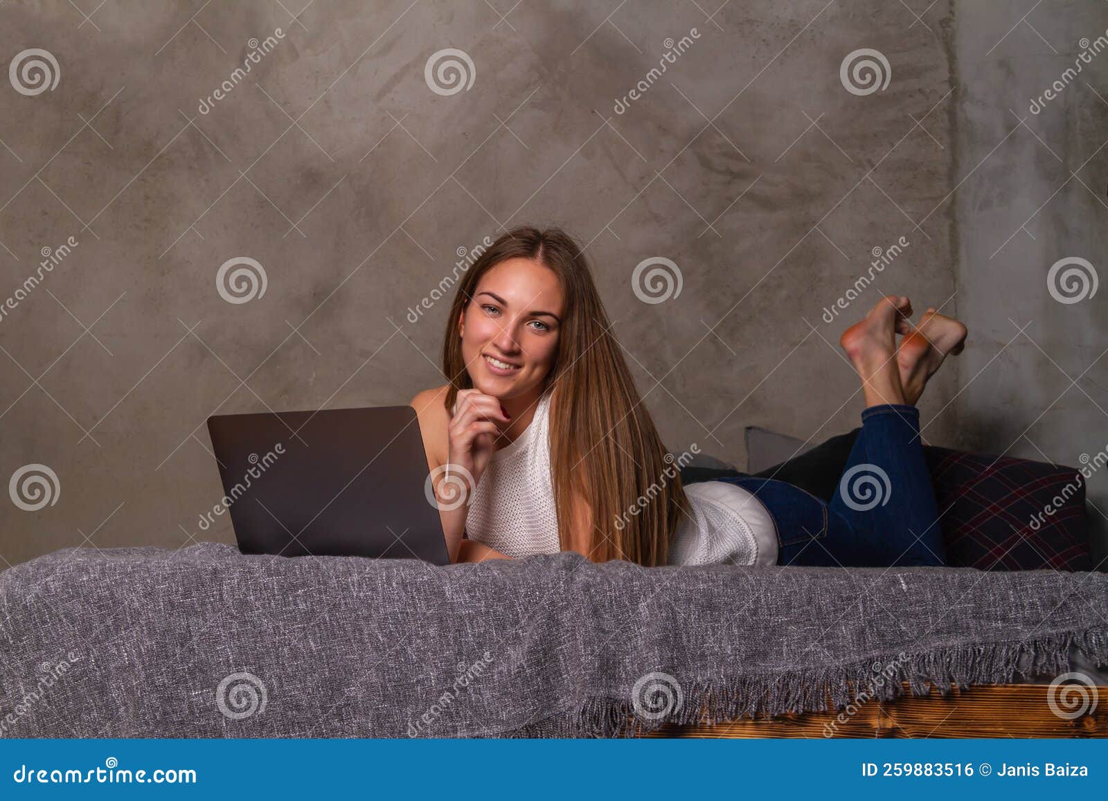 Smiling Woman Lying On Her Stomach In Bed With Laptop In Front Of Her