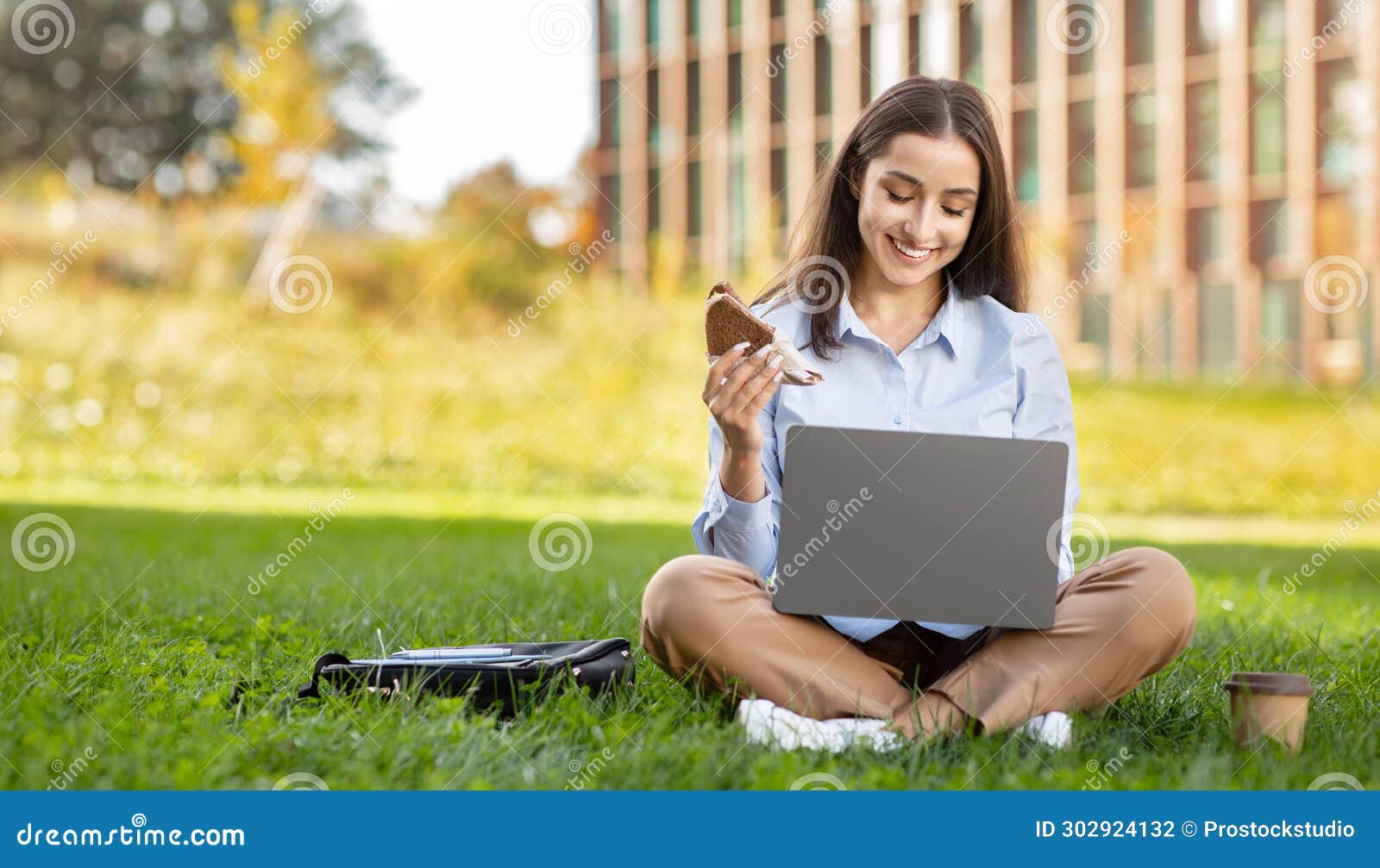 Smiling Young Professional Woman Multitasking With A Laptop On Her Lap Enjoying A Sandwich