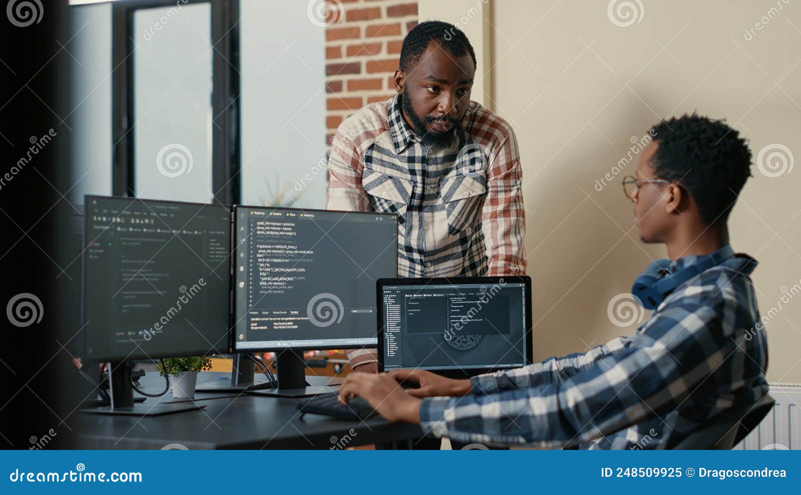 Software Developer Sitting At Desk With Multiple Screens And Laptop Running Code Talking With