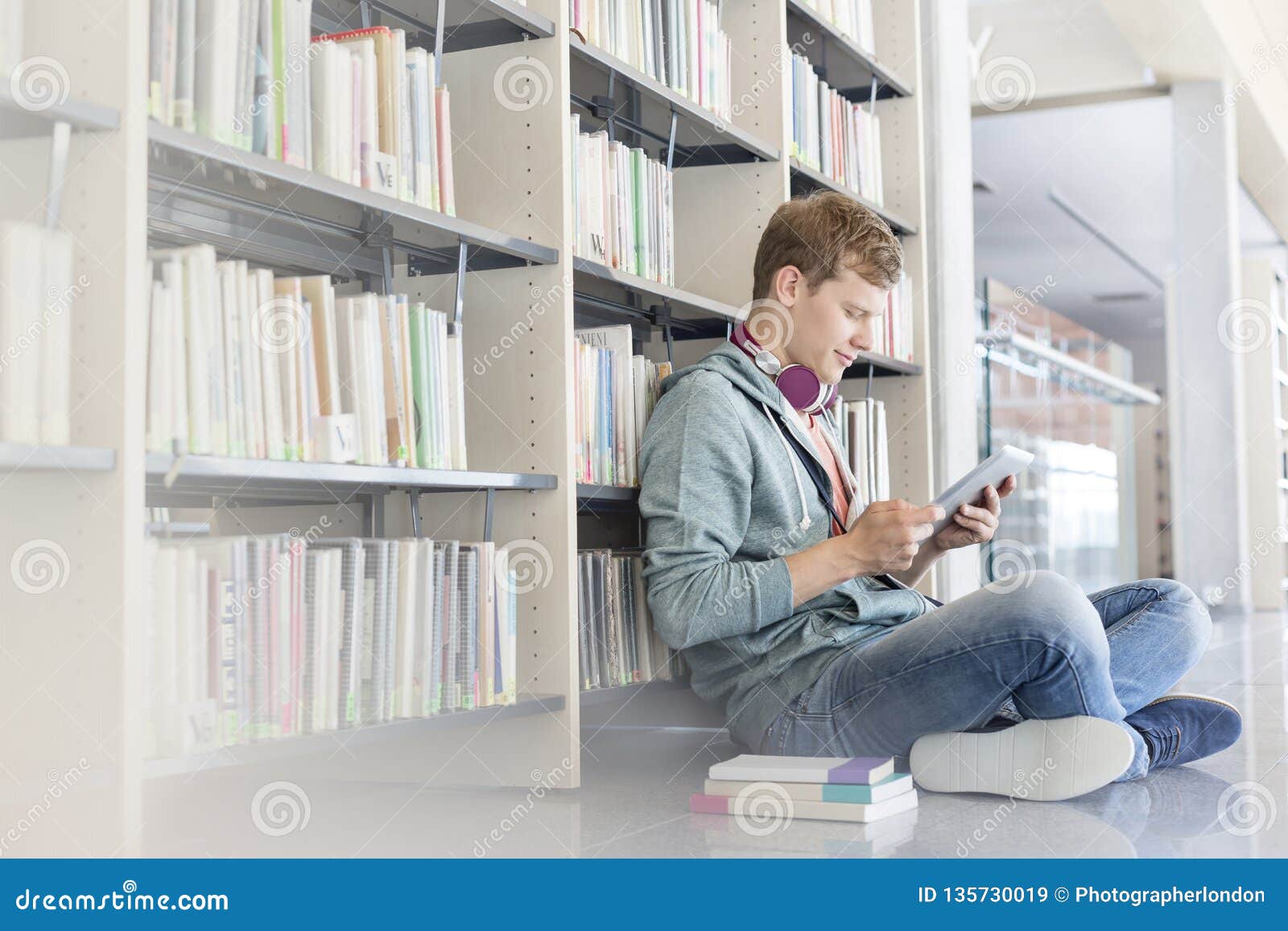 Student Using Digital Tablet Against Bookshelf while Sitting on Floor ...