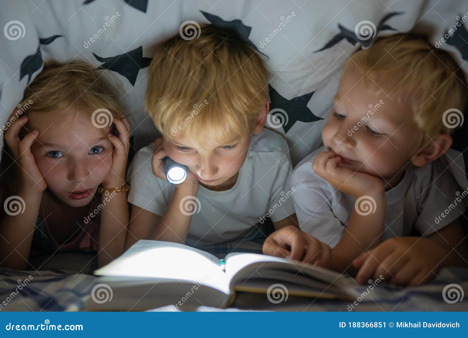 Three Young Children are Reading a Book with a Flashlight Under the ...