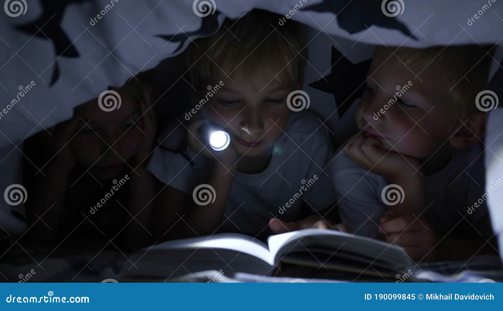 Three Young Children are Reading a Book with a Flashlight Under the ...