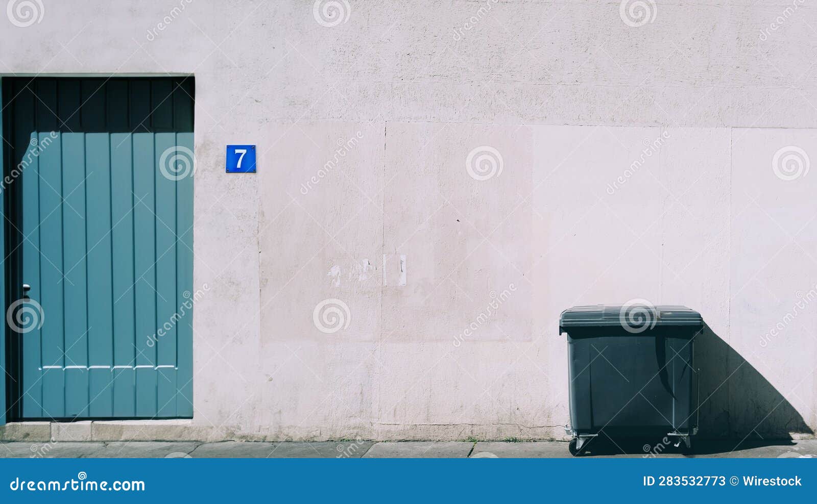 Trash Bin in Front of a Building Wall with a Blue Door Illuminated by ...