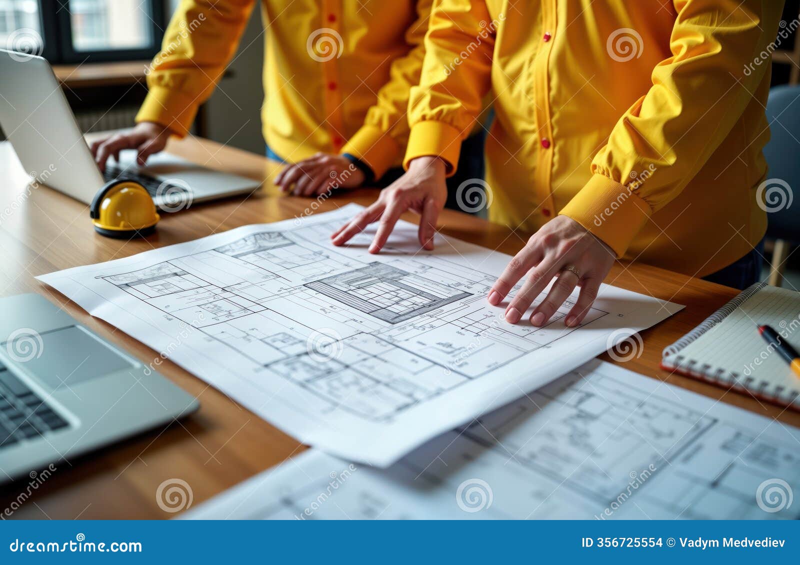 Two Engineers in Yellow Shirts Review Architectural Plans at Desk. Look ...