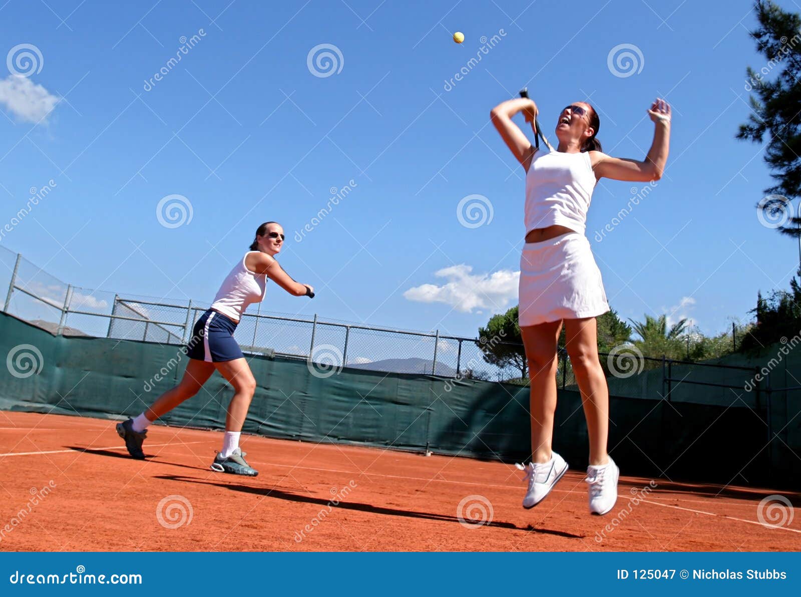 Two Female Tennis Players Playing Doubles In The Sun One Is Leaping