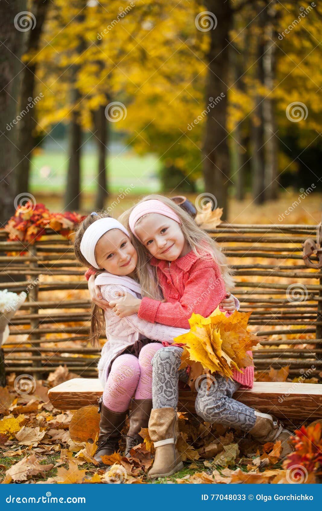 Two Girls In Autumn Park Sitting On Wooden Bench Near A Fence Stock