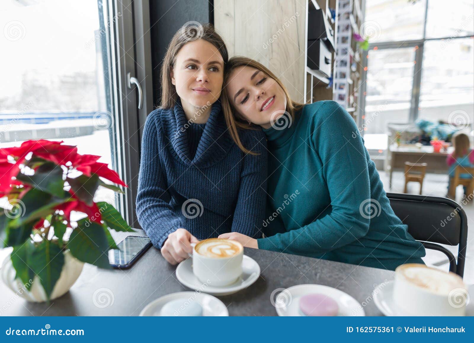 Two Laughing Embracing Young Women Sitting At Table In Coffee Shop