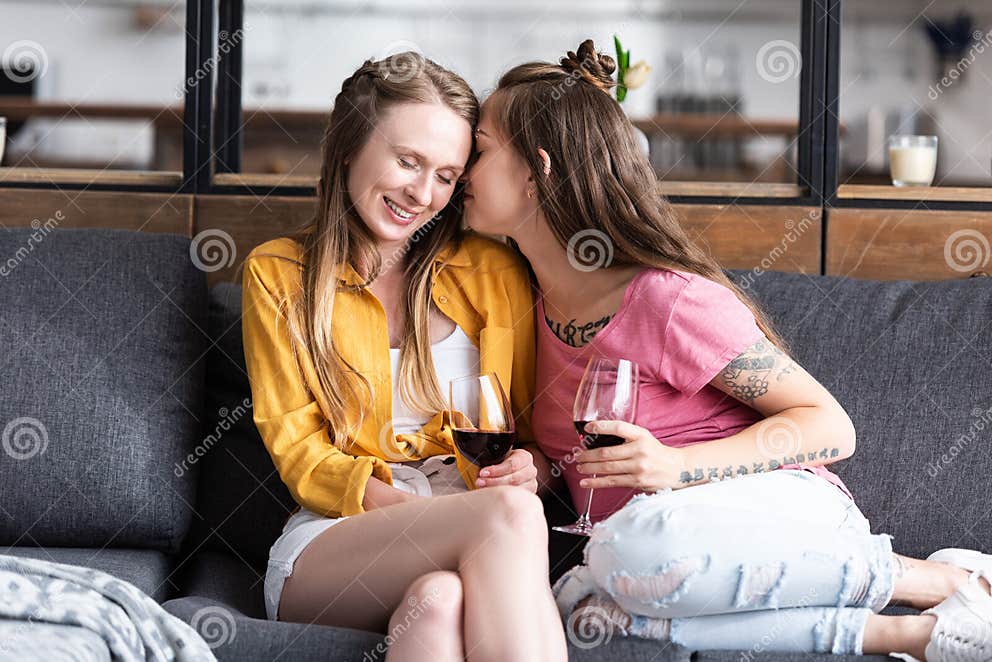 Two Lesbians Holding Wine Glasses And Kissing While Sitting On Sofa In