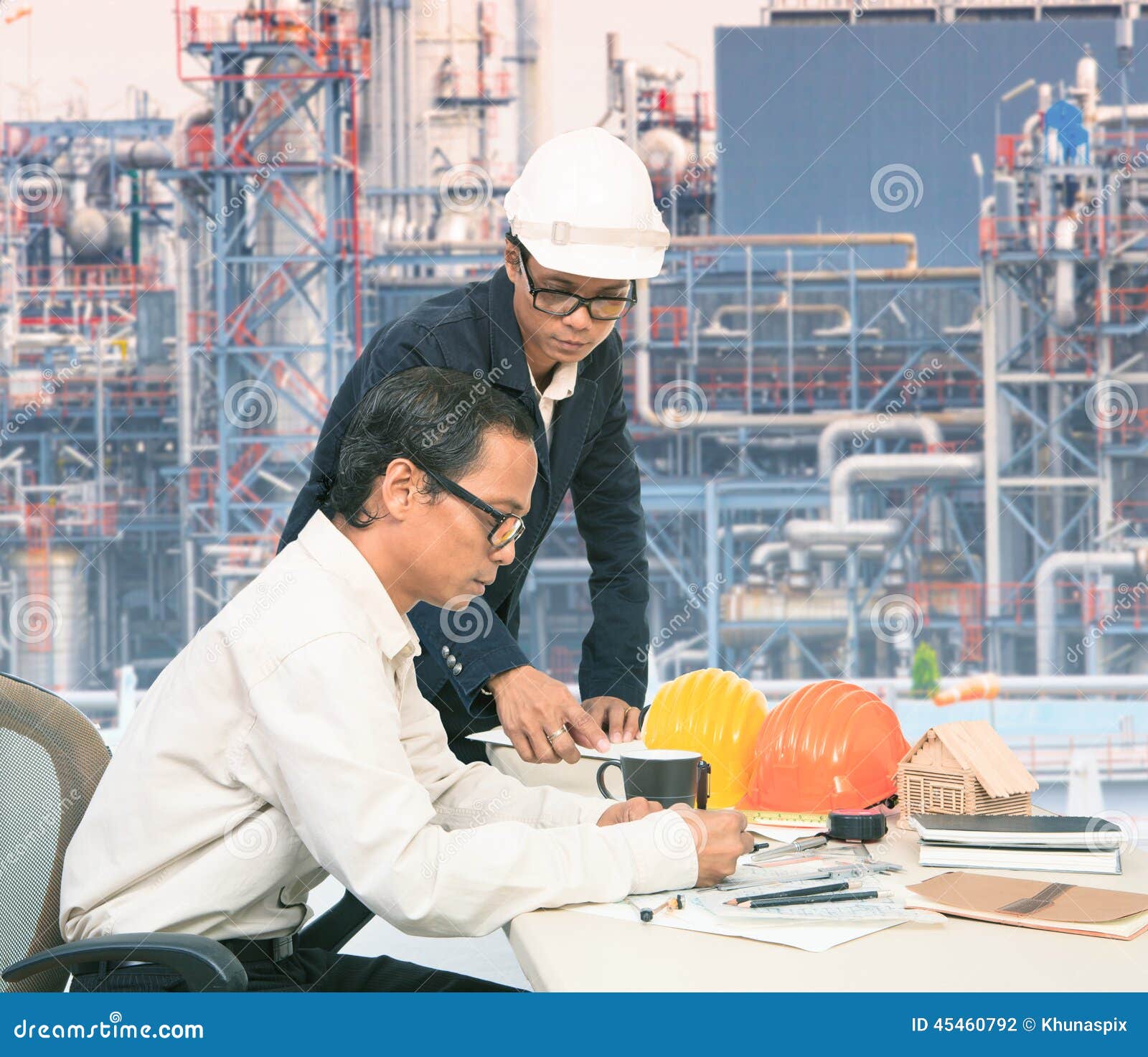 Two of Same Engineer Working on Table Against Exterior of Oil Refinery ...