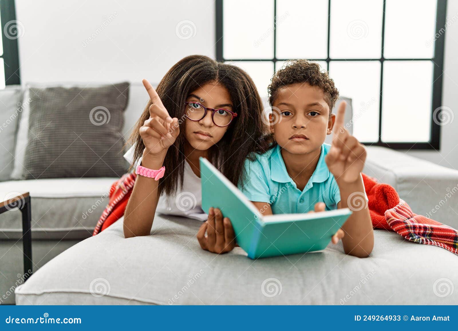 Two Siblings Lying on the Sofa Reading a Book Pointing with Finger Up ...