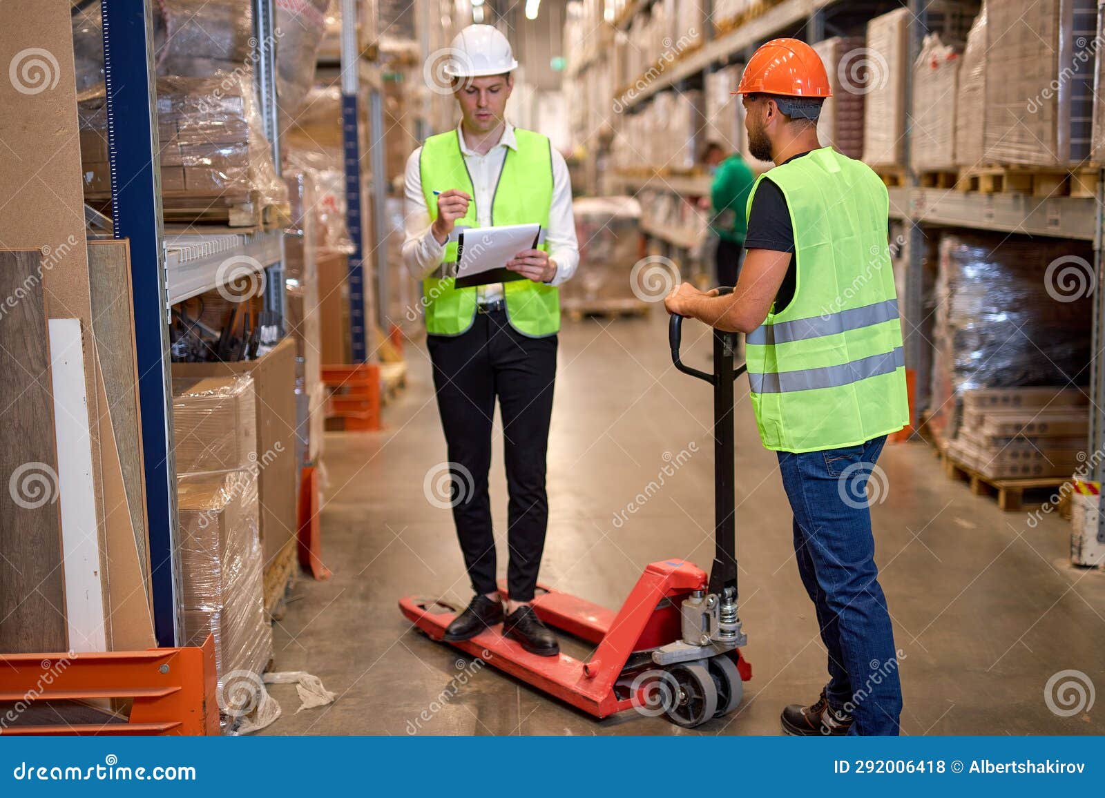 Two Warehouse Workers in Safety Vasts and Helmets Talking Together at ...