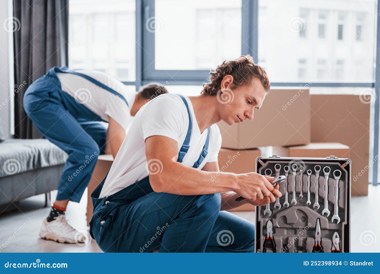 Uses Tools from Case. Two Young Movers in Blue Uniform Working Indoors ...