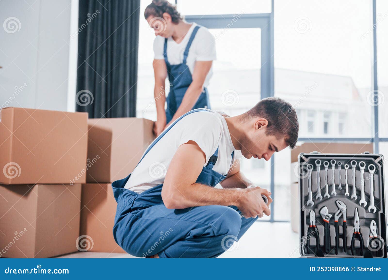 Uses Tools from Case. Two Young Movers in Blue Uniform Working Indoors ...