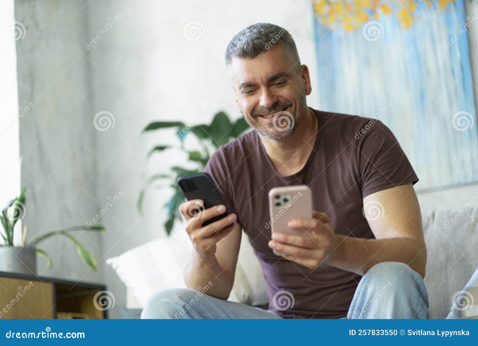 Using Two Modern Smartphones Sitting on the Couch Handsome Grey Haired ...