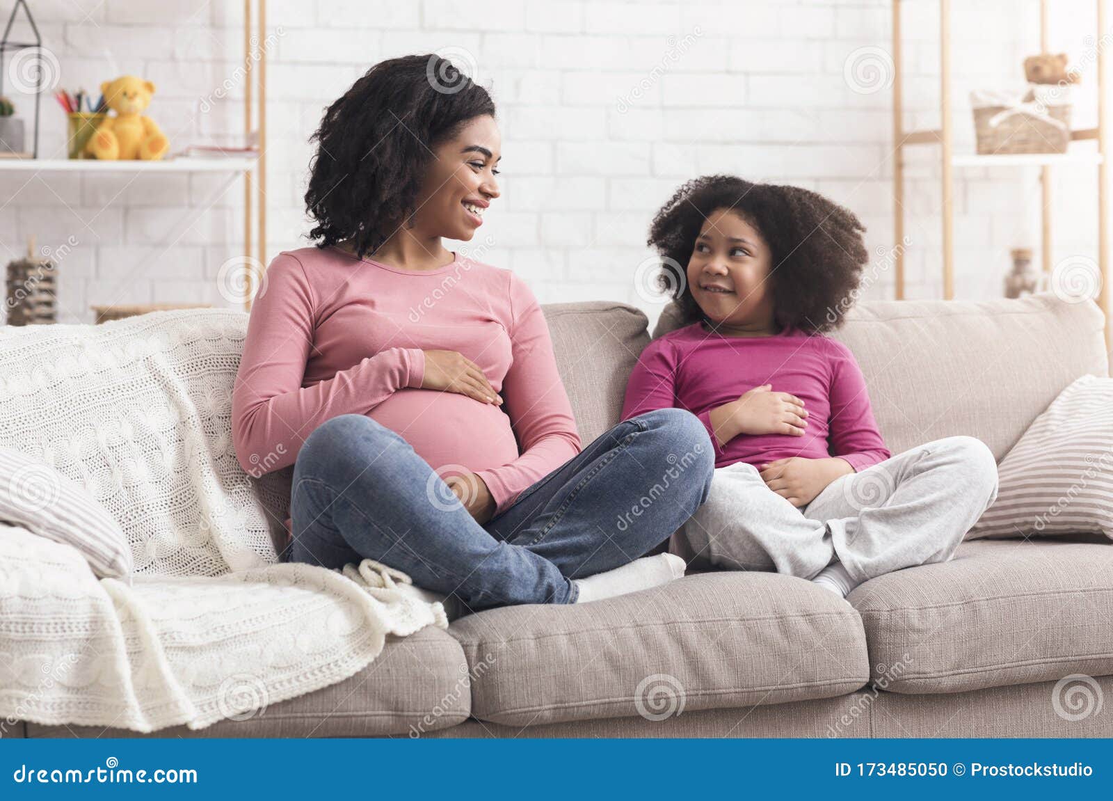Pregnant Afro Mom And Her Daughter Touching Bellies Sitting On Couch