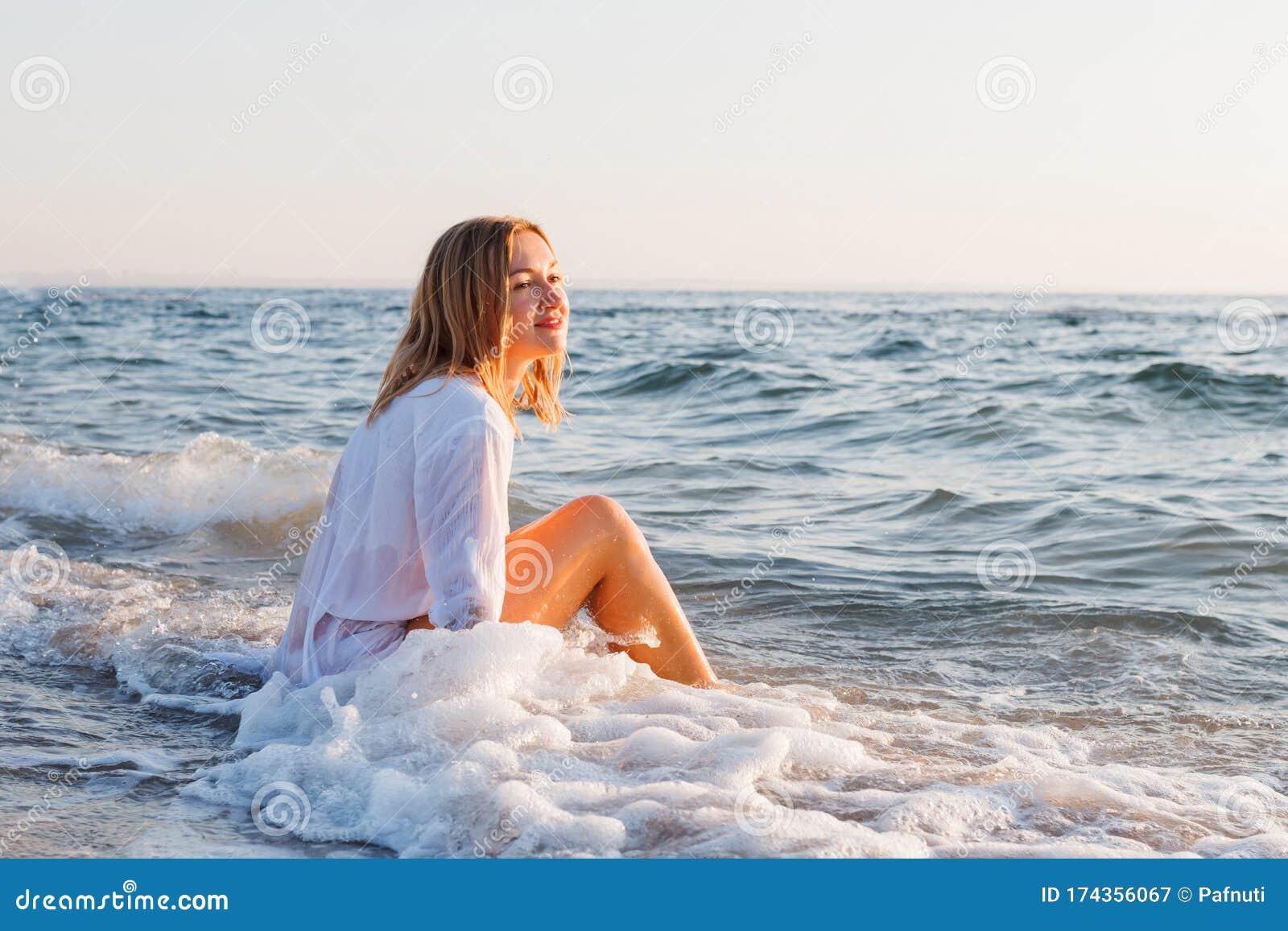 Wet Blonde Girl In White Shirt Sitting In The Water On The Sea Beach