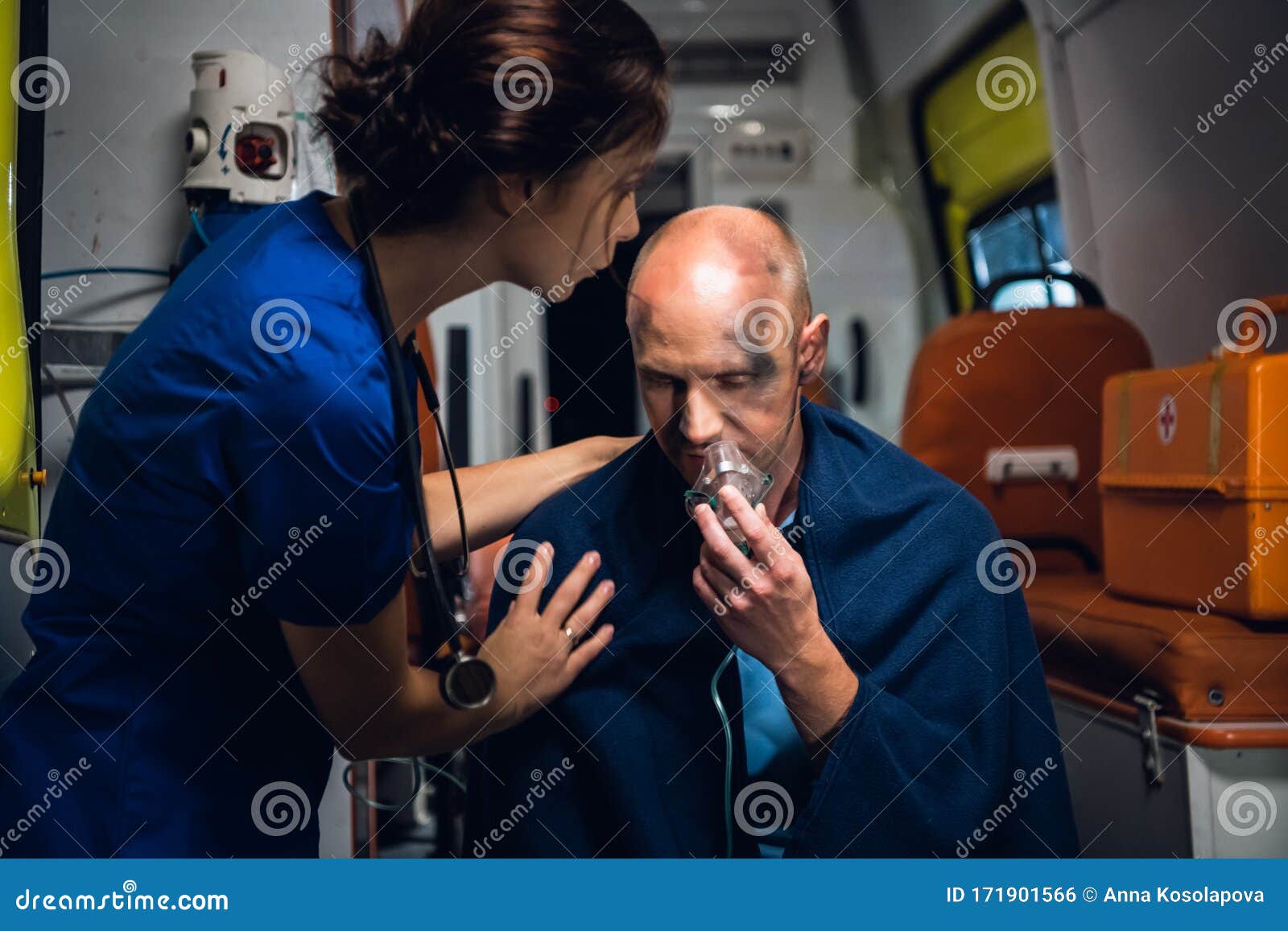 Woman In Blue Medical Uniform Stands With Man Who Sits In Oxygen Mask