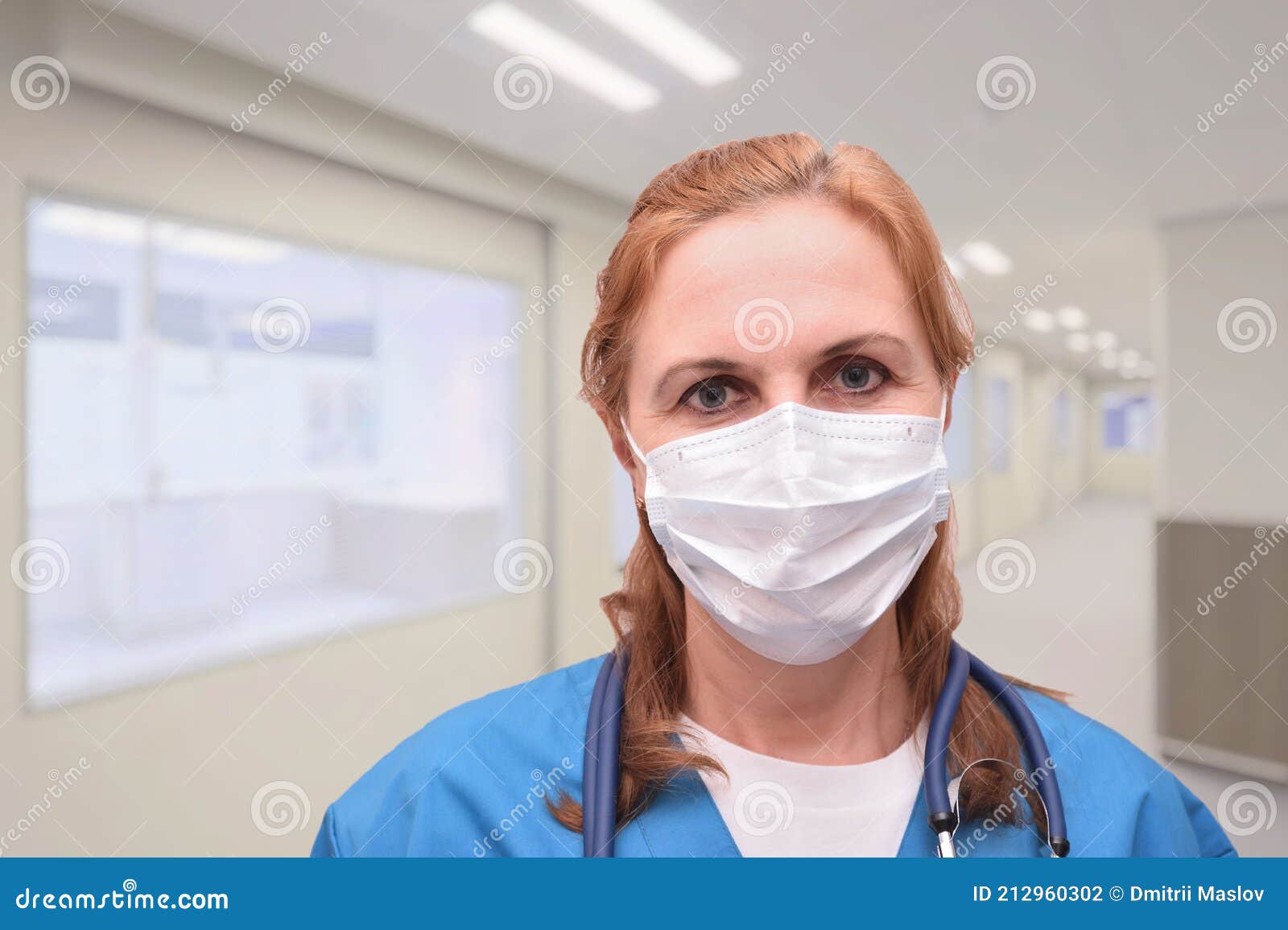 Female Doctor In A Blue Medical Suit And Mask Stands In The Corridor Of