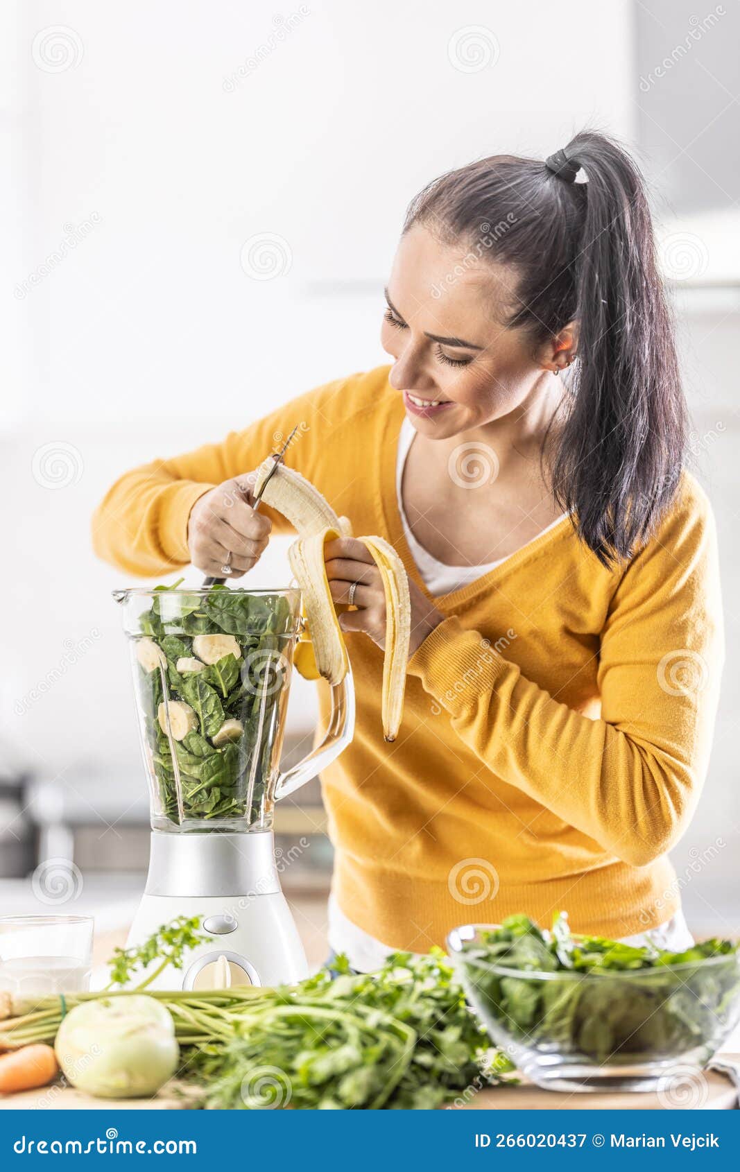 A Woman In The Kitchen Cuts A Banana In A Blender Together With Spinach