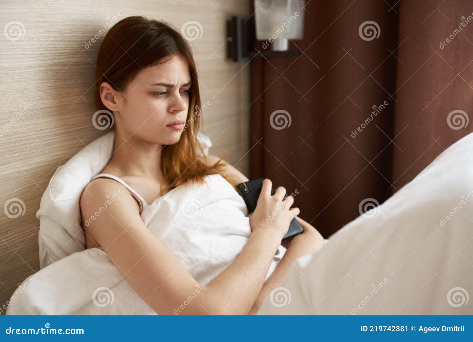 Woman Lies In Bed Under The Covers With A Mobile Phone In Her Hand