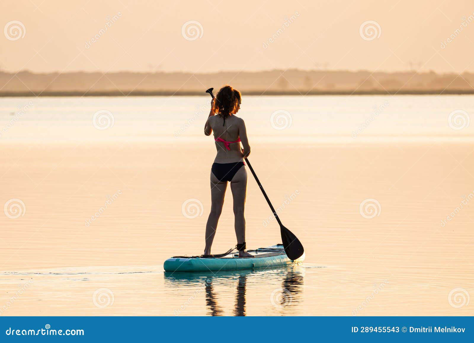 A Woman In Mohawk Shorts Stands On A Sup Board At Sunset In A Lake