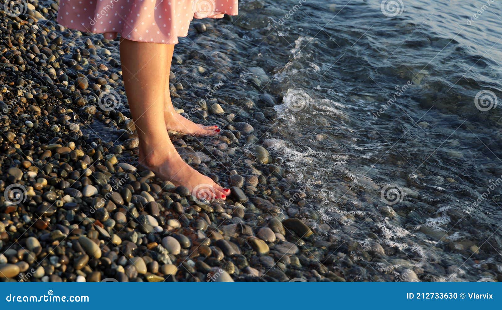 Woman`s Legs Stand Barefoot On Seaside Pebble Shore In Light Surf Stock
