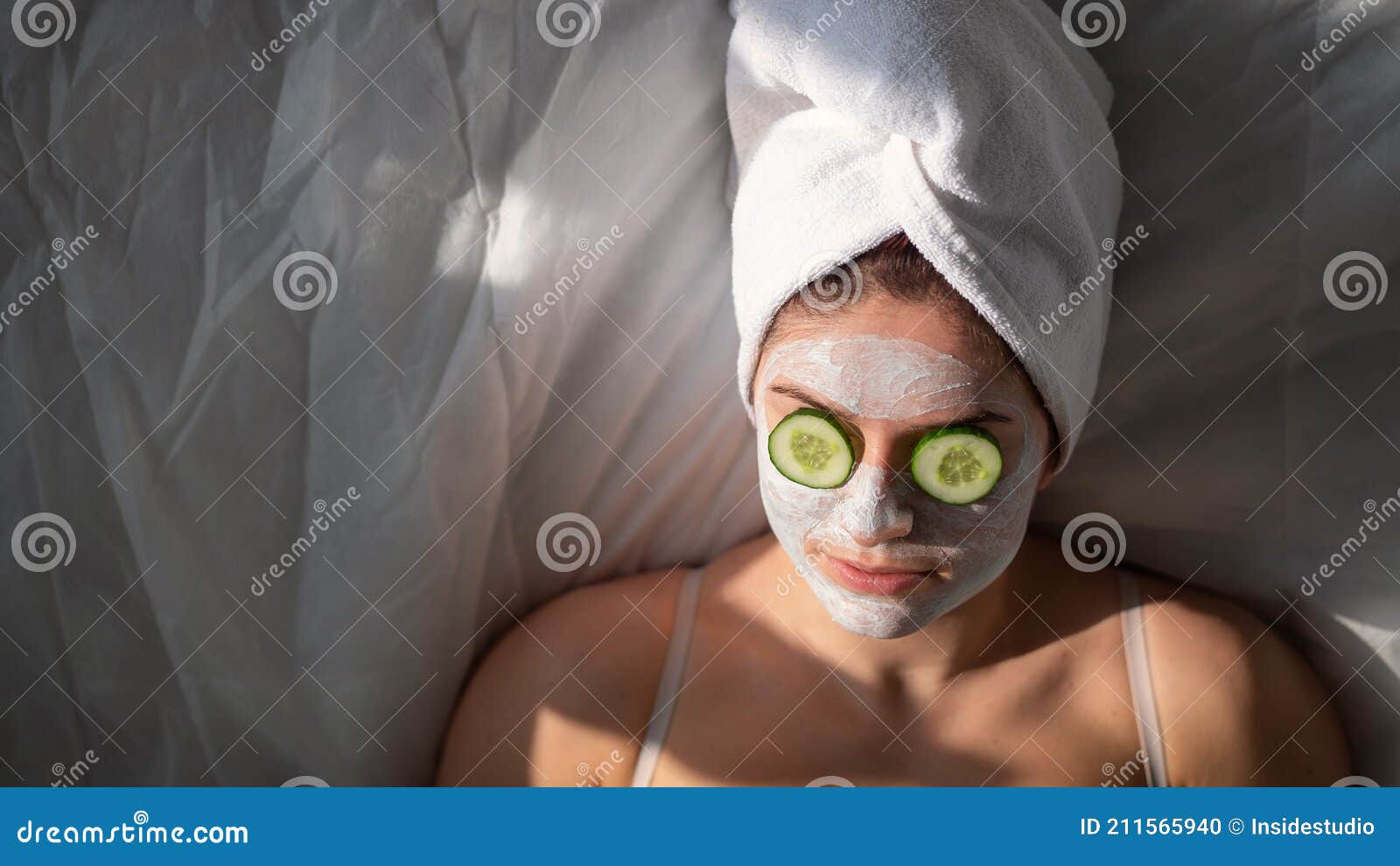 A Woman With A Towel On Her Hair And In A Clay Face Mask And Cucumbers
