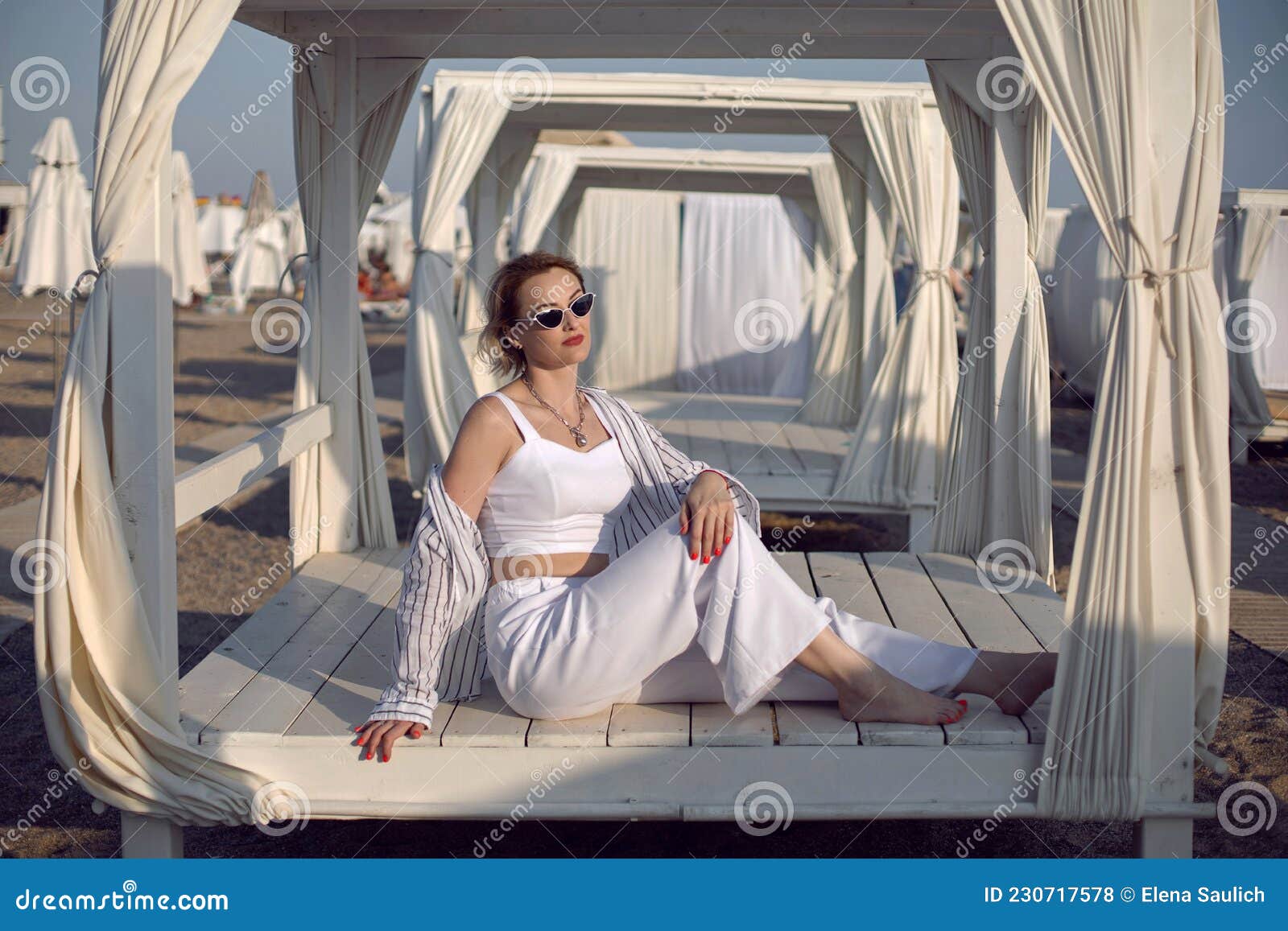 Woman In White Clothes And Sunglasses Is Sit On The Beach Near Wooden