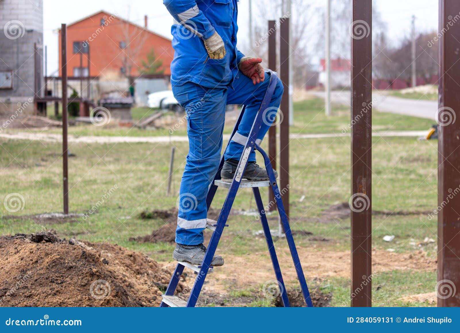 Worker in Blue Overalls Stands on a Metal Ladder in the Yard Stock ...