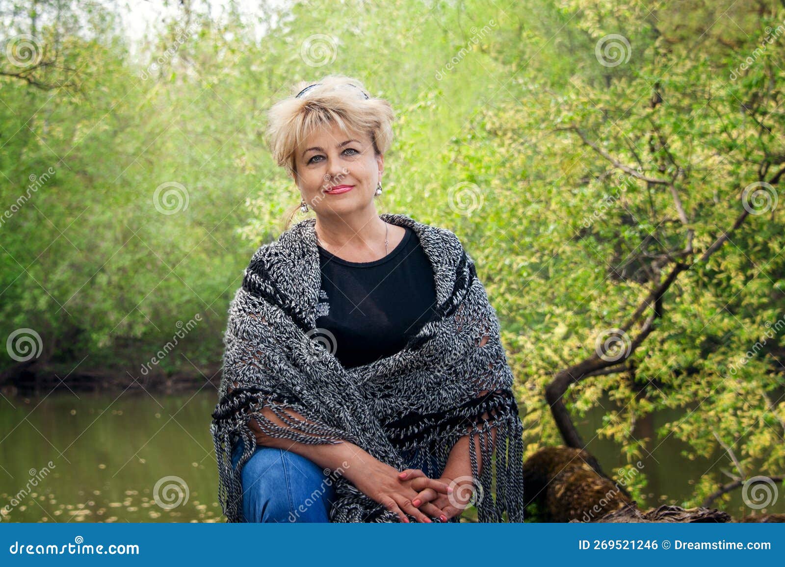 55 60 Year Old Woman In A Knitted Shawl Is Sitting On The Riverbank