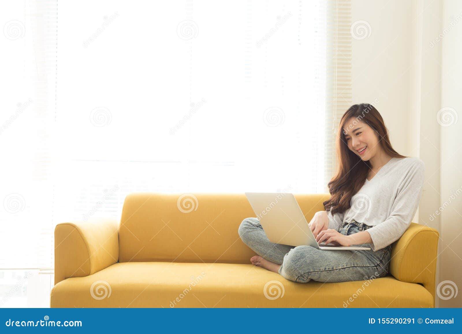 Young Asian Woman Sitting With Laptop On Yellow Couch In Home Office