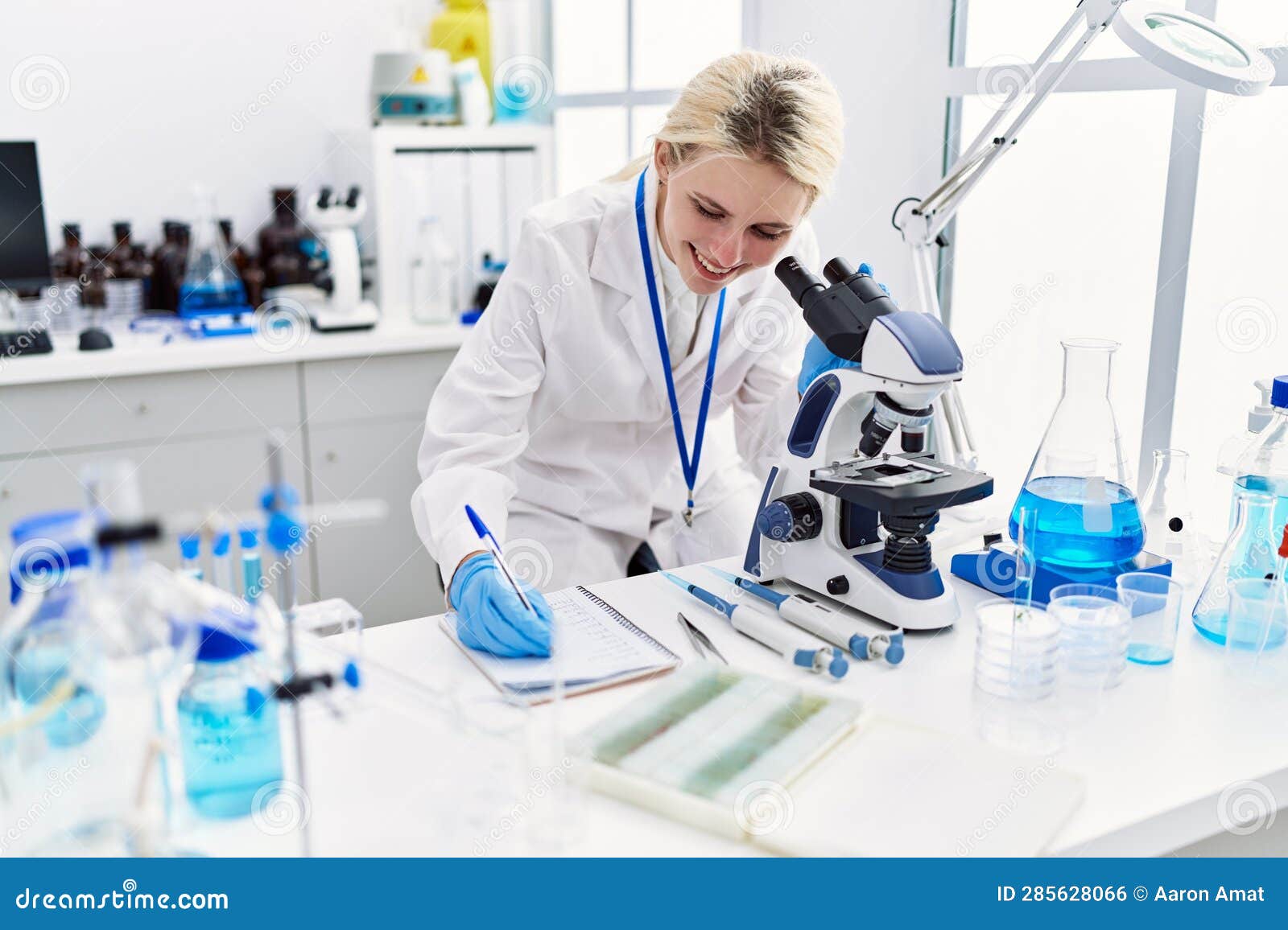 Young Blonde Woman Scientist Using Microscope Taking Notes at ...