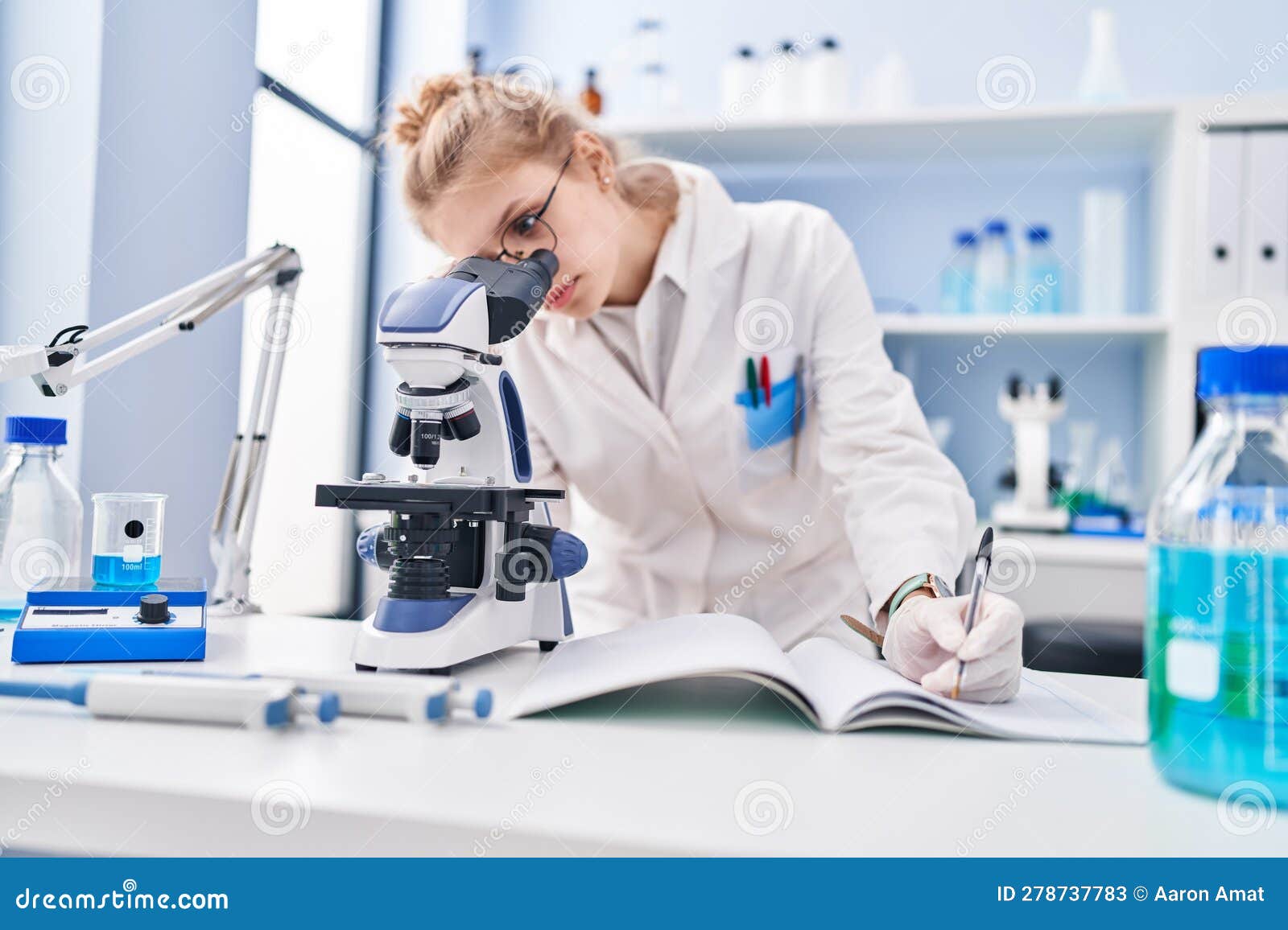 Young Blonde Woman Scientist Using Microscope Writing Report at ...