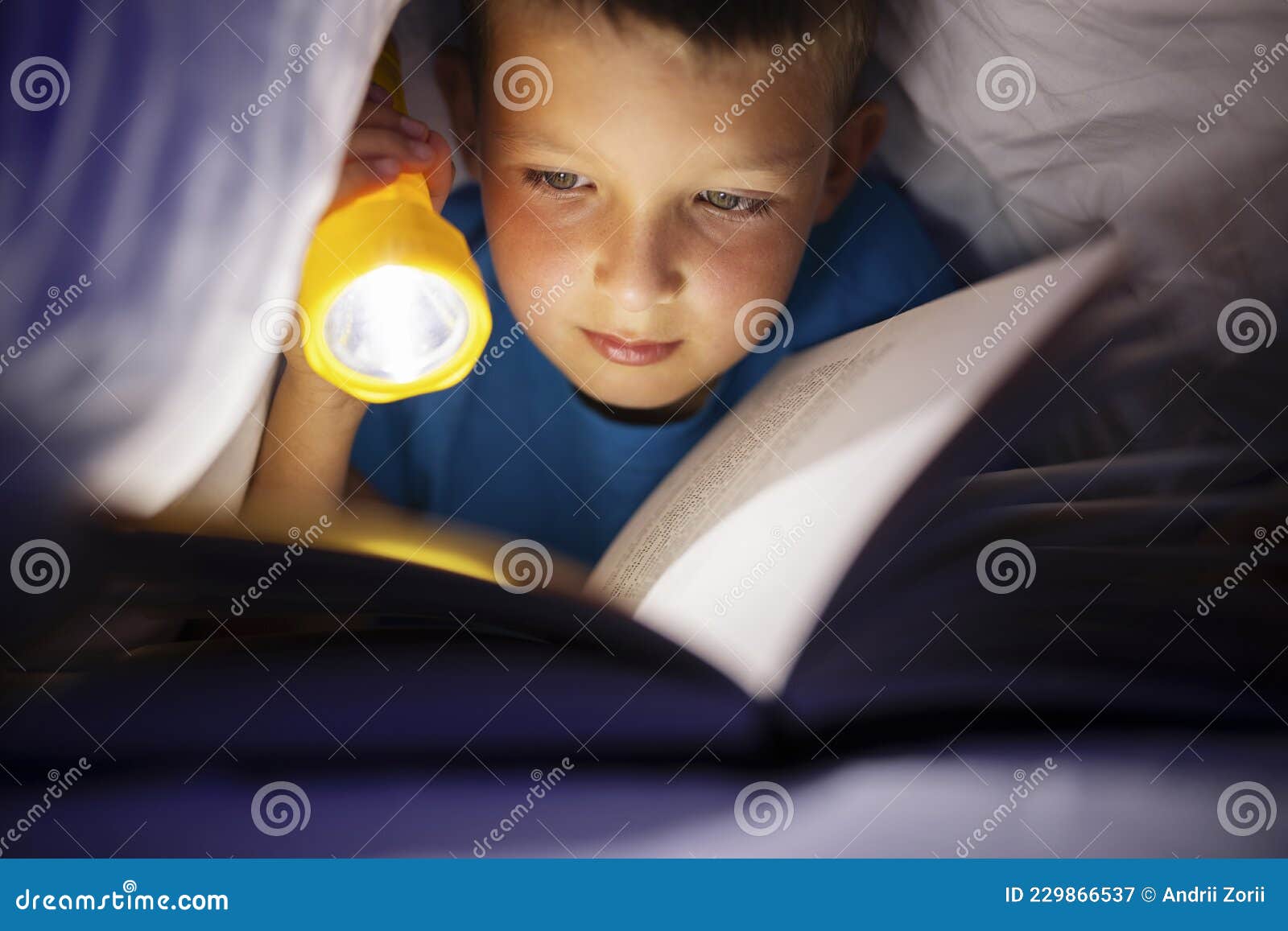 A Young Boy Reading a Book Under the Covers with a Flashlight at Dark ...
