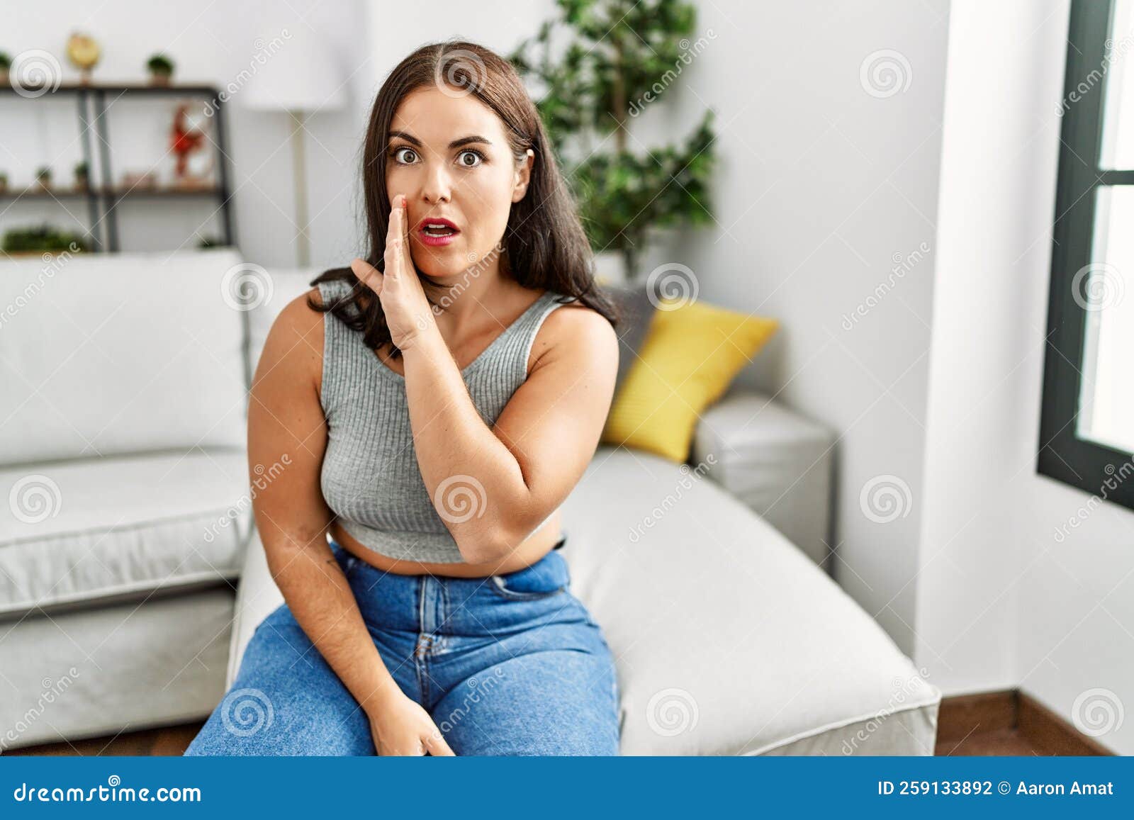 Young Brunette Woman Sitting On The Sofa At Home Hand On Mouth Telling