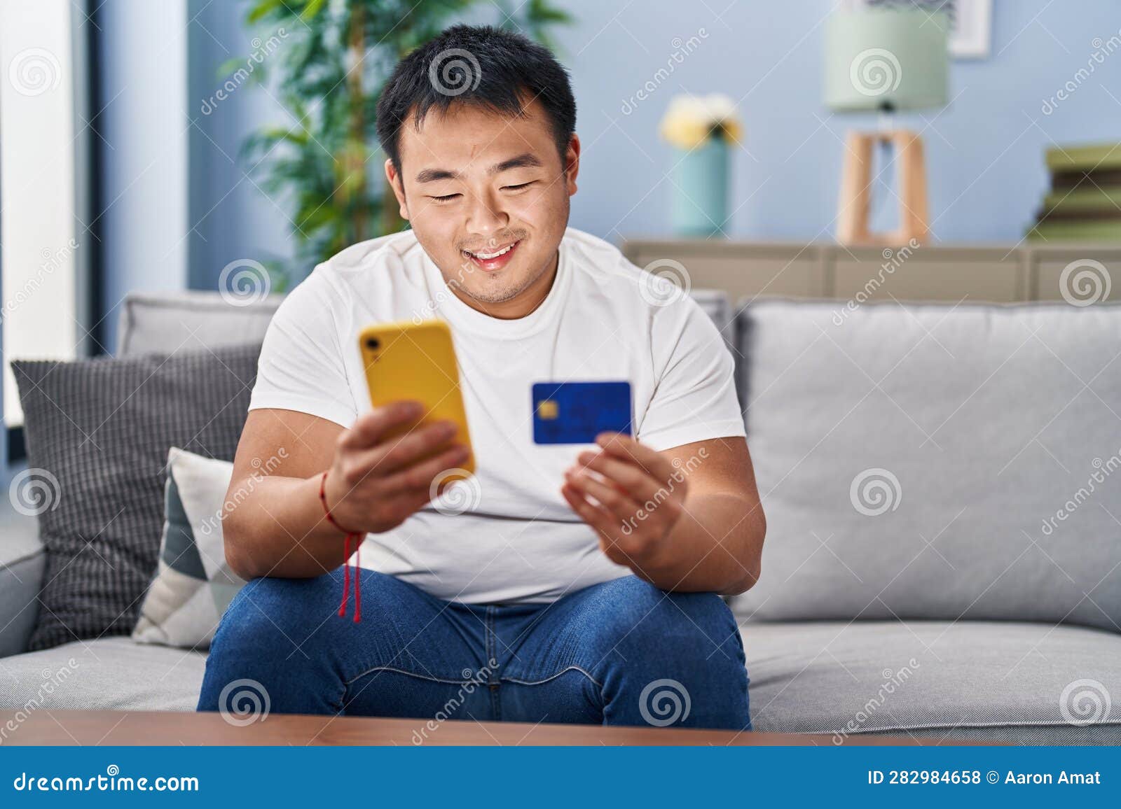 Young Chinese Man Using Smartphone and Credit Card Sitting on Sofa at ...