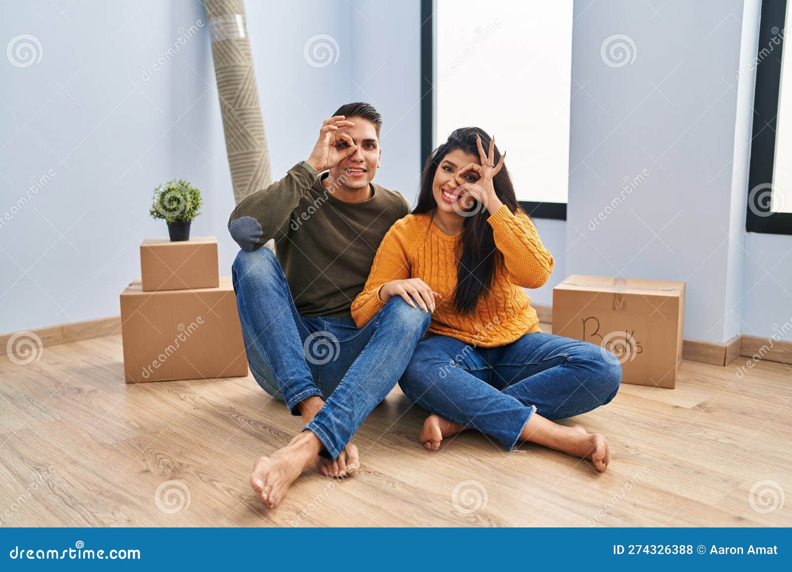 Young Couple Sitting On The Floor At New Home Doing Ok Gesture With