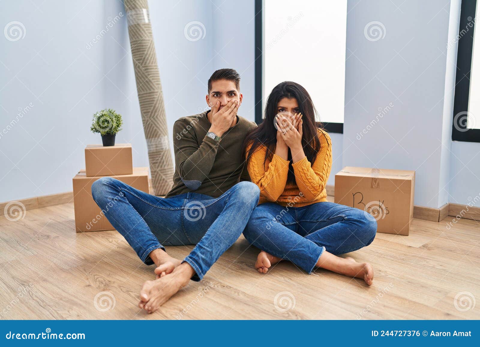 Young Couple Sitting On The Floor At New Home Shocked Covering Mouth