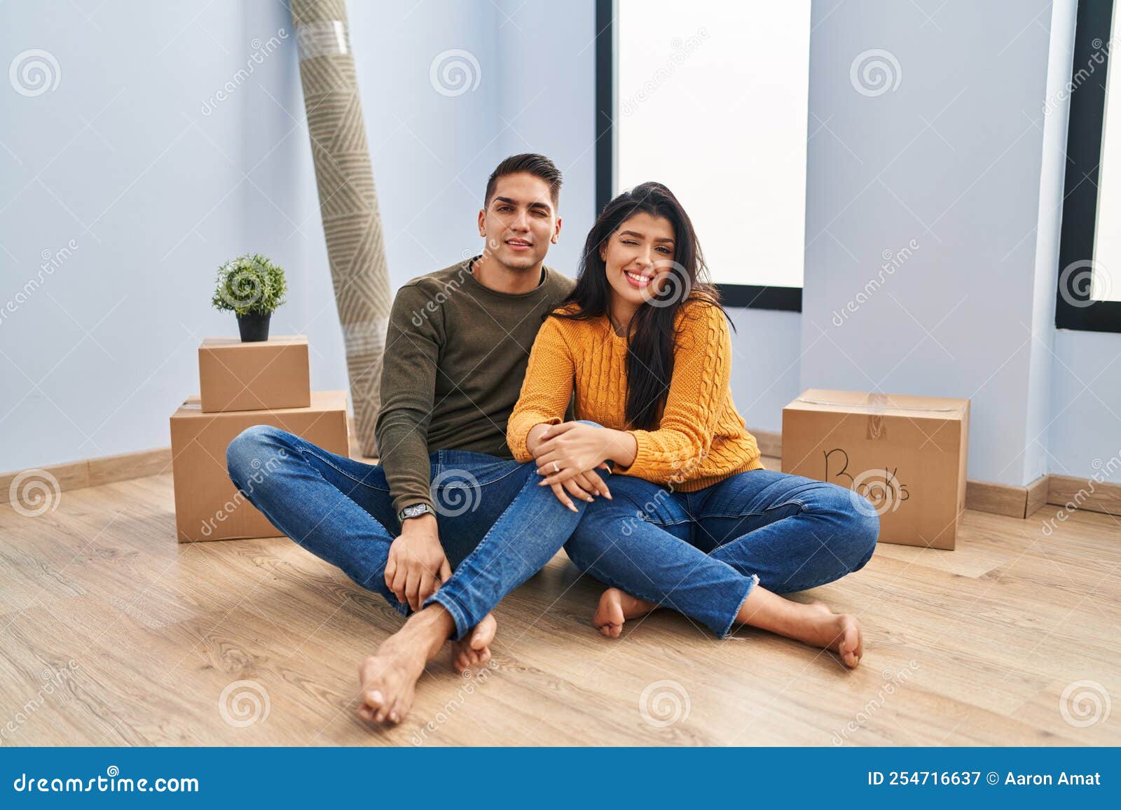 Young Couple Sitting On The Floor At New Home Winking Looking At The