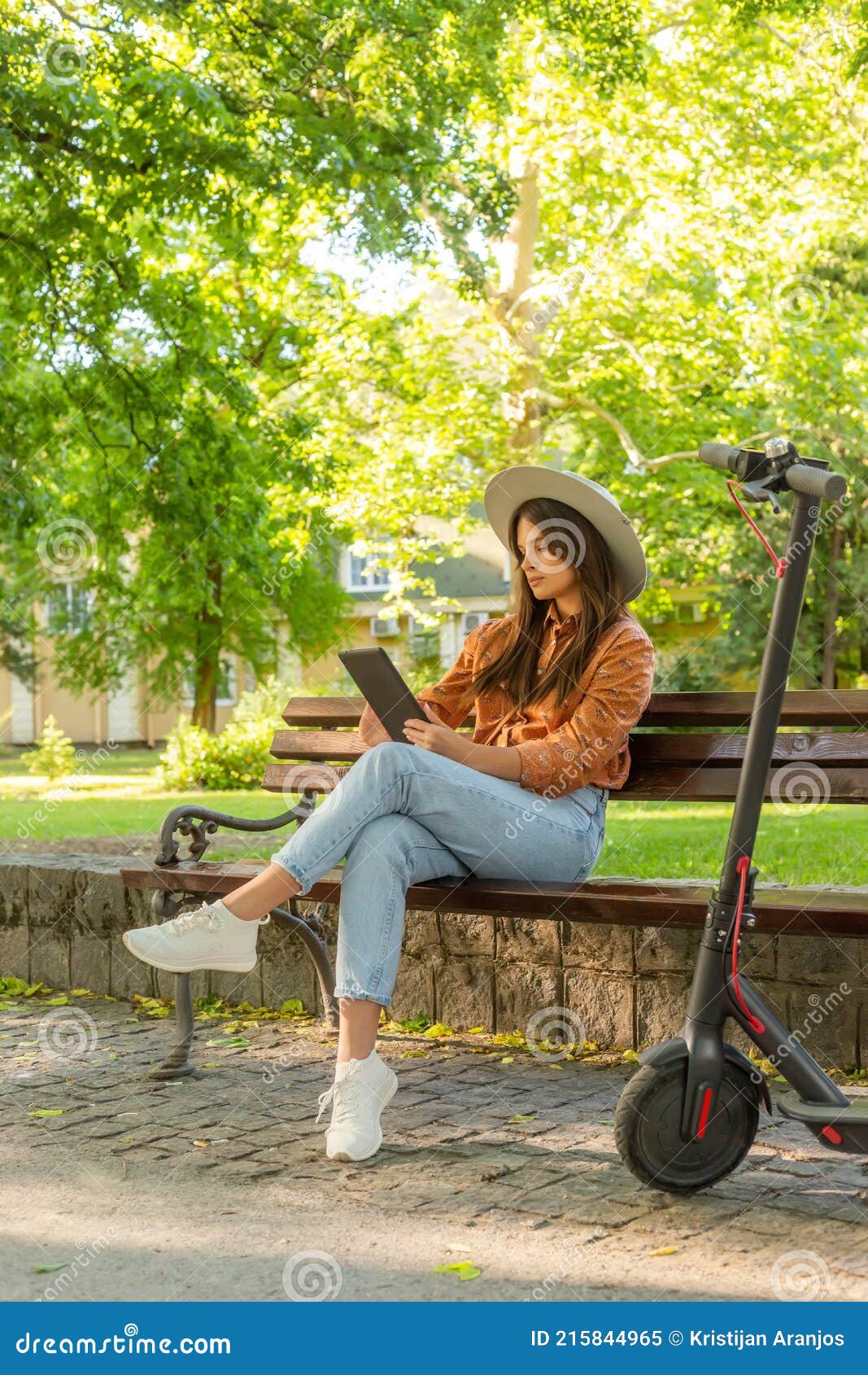 A Young Girl Is Sitting On A Park Bench With The Tablet In Her Hands