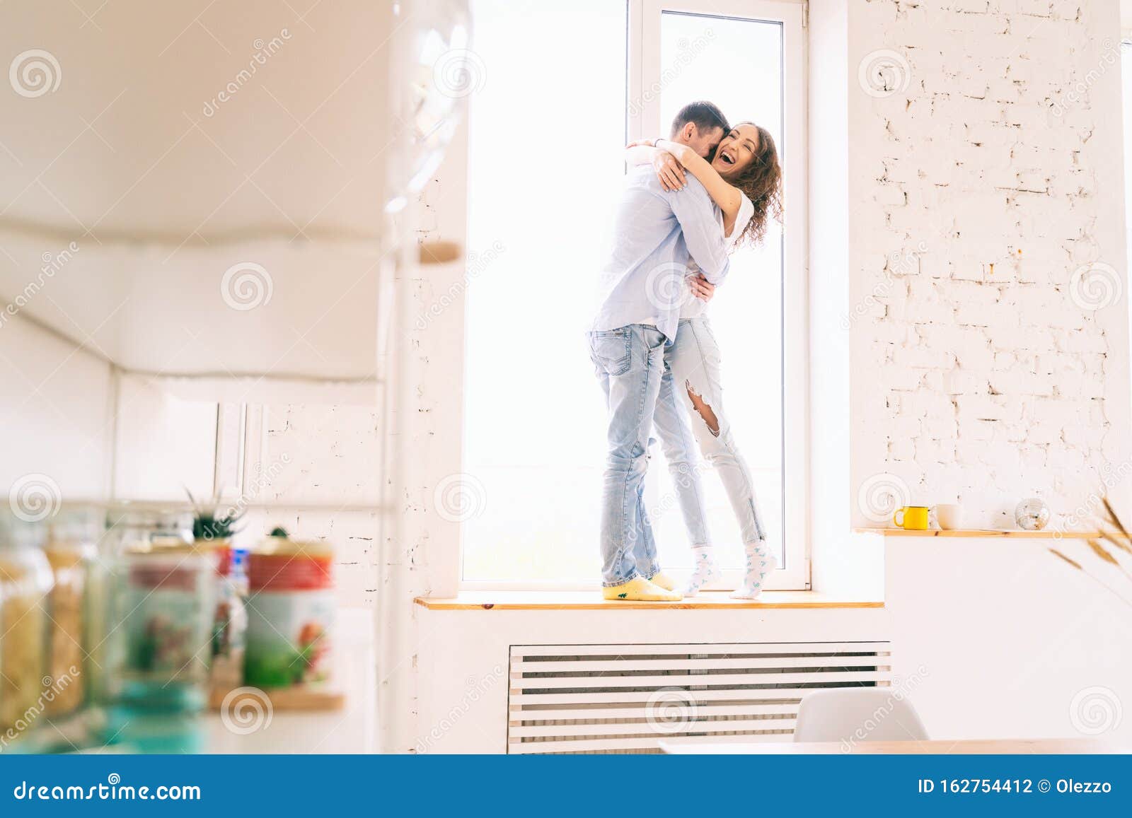 Young Happy Couple Hugging On The Windowsill In Their Apartment