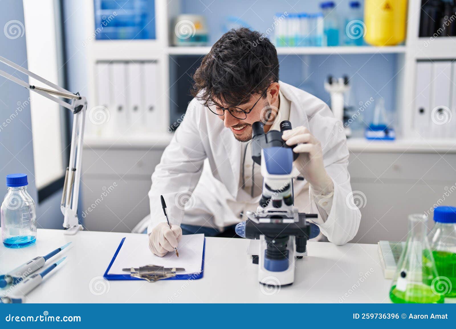Young Hispanic Man Scientist Writing on Document Using Microscope at ...