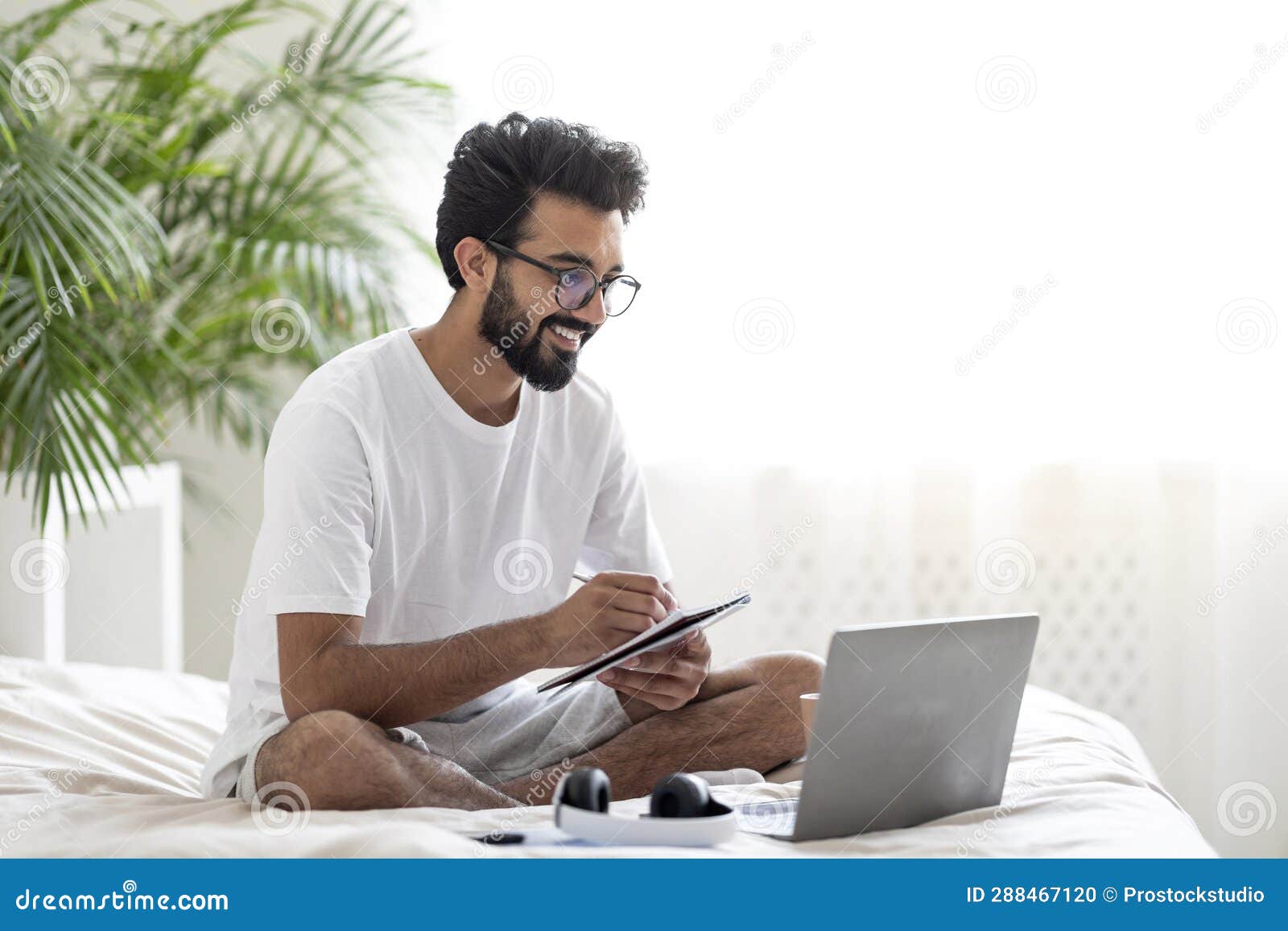Young Indian Man Sitting in Bed Using Laptop and Writing in Notepad ...